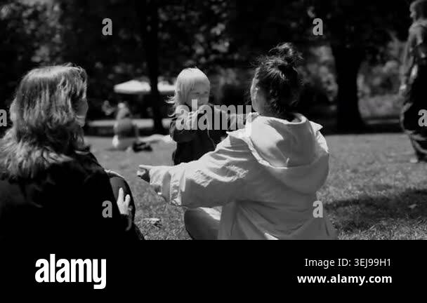 Park scene caregiver holds out toddler sitting on grass under trees ...