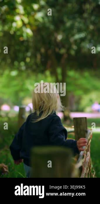 White toddler tracing fence rope near park bench, turning back to look ...