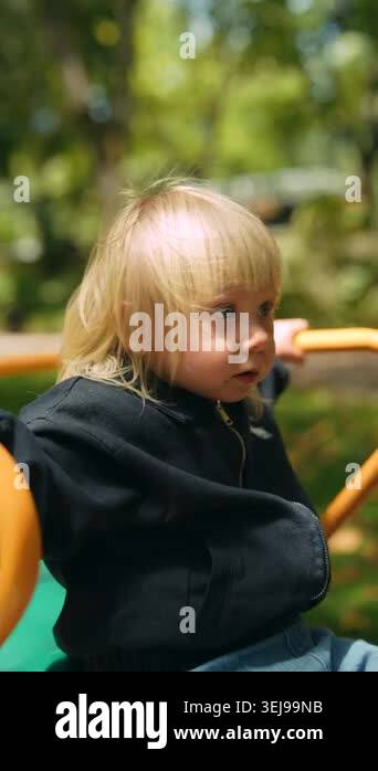 White toddler on playground spinner holding yellow handle, leafy park ...