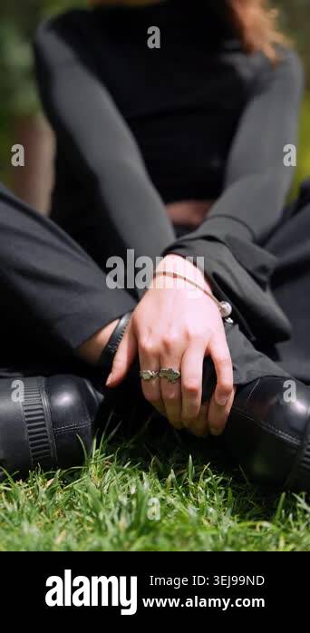 Woman sitting crosslegged on grass, black outfit with boots and rings ...