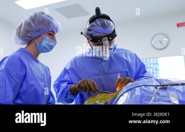 Male neurosurgeon in surgical loupes applies metal tools. Female nurse ...