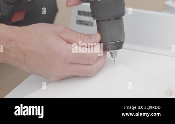 Hands hold cordless drill making small hole in white furniture panel ...