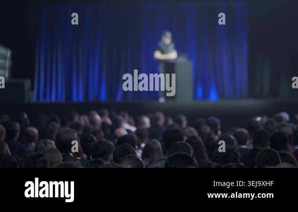 Large audience watching lone speaker deliver presentation on dimly lit ...