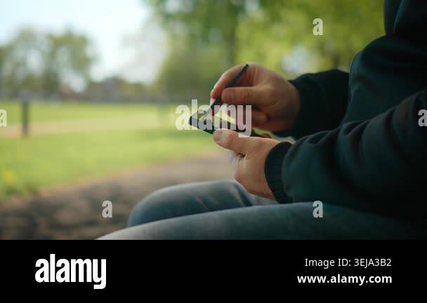 Stylus tapping smartphone in hands while sitting in park with green ...