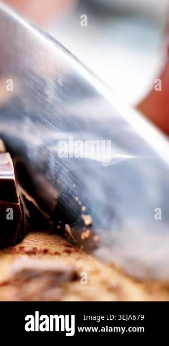 close up of chef knife chopping dark chocolate bar on wooden board ...