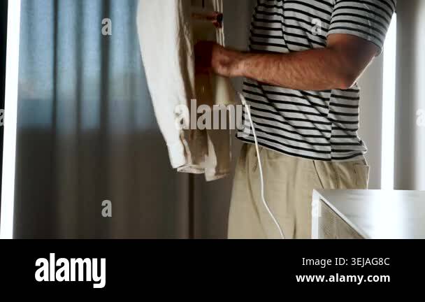 Man holds a light beige button up shirt in an indoor room. He wears a ...