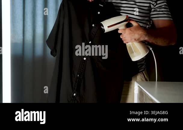 A person in a striped shirt steams a black button-up shirt on a hanger ...
