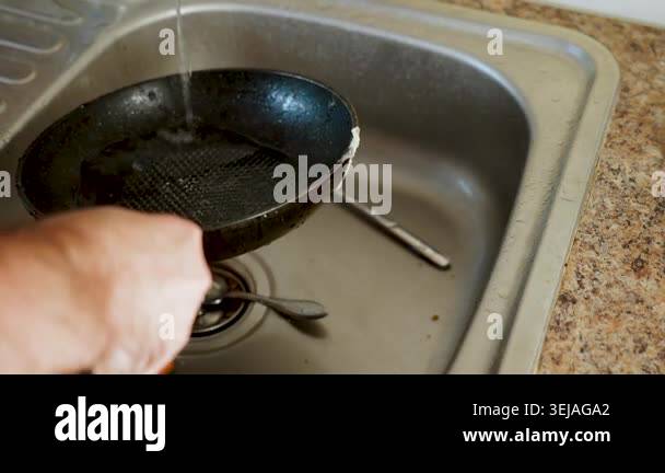 A person's hand cleans a dirty frying pan under a stream of water in a ...