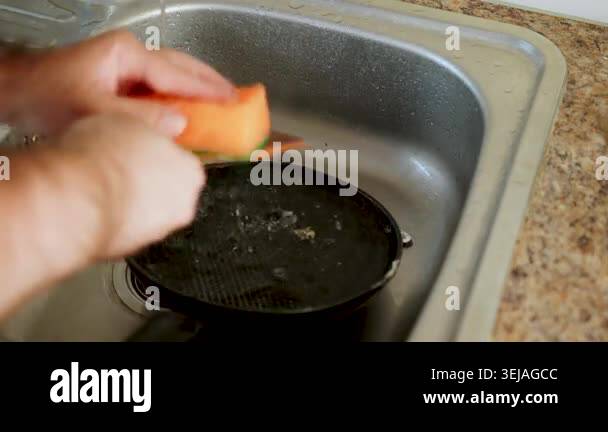 Hands scrub a dirty frying pan and a fork with an orange sponge in a ...