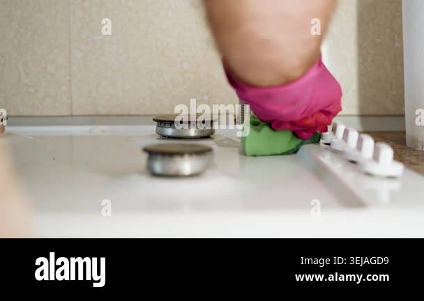 A person wearing a pink glove scrubs a white gas stove with a green ...