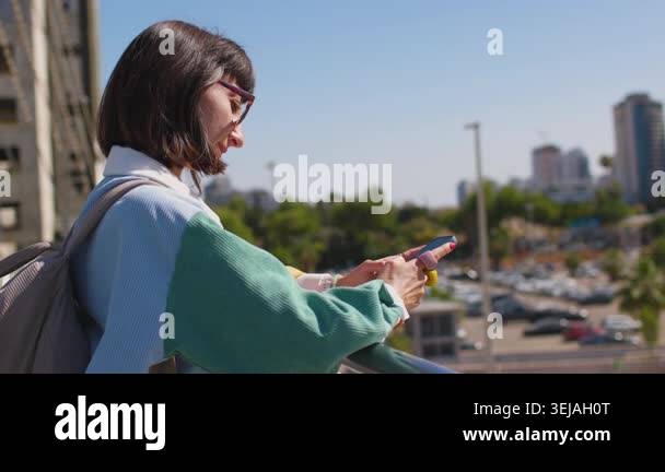 A young woman texting on the phone, leaning on a railing, wearing ...