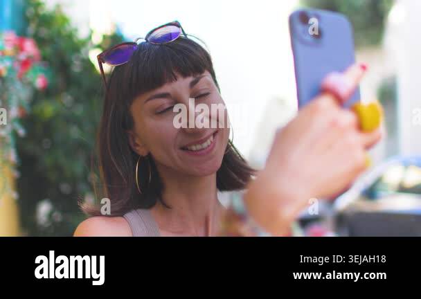 A smiling woman takes a photo using her phone on a sunny city street ...