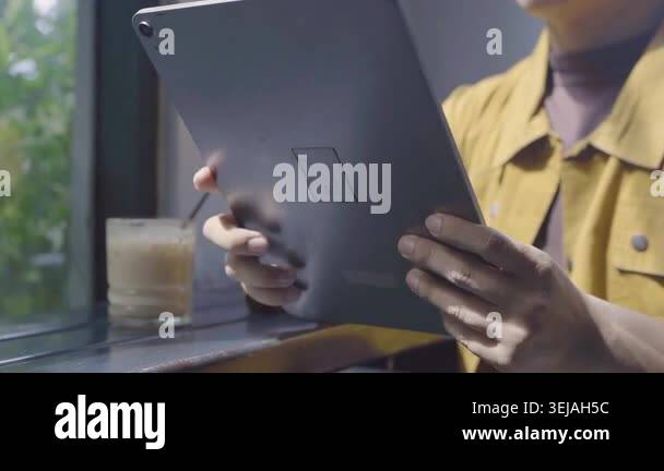 Asian hands scrolling tablet near window, green foliage outside glass ...