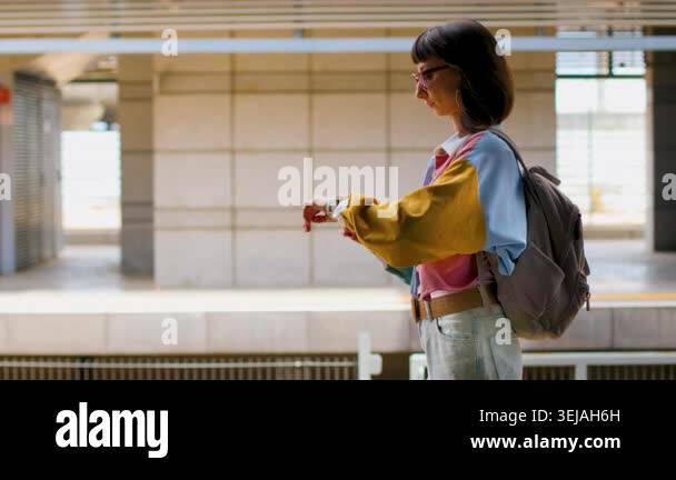 A smiling woman waits for a train and uses her smartwatch at a train ...