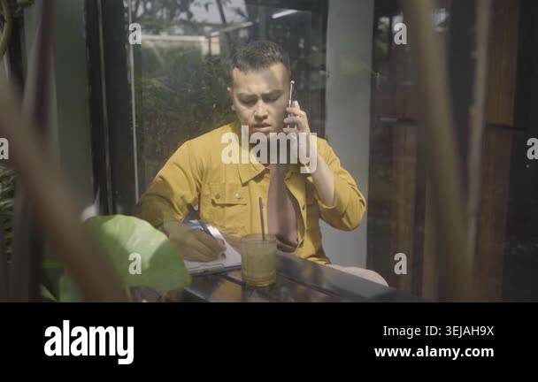 Asian man on phone at cafe, writing notes in notebook, sipping coffee ...