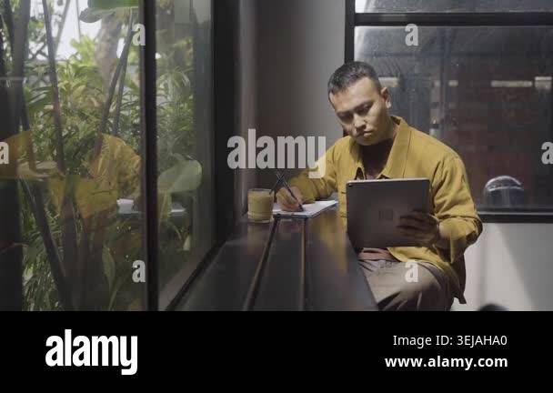 Asian man journaling by window seat, soft natural light, indoor plants ...
