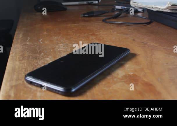 A smartphone rests on a wooden desk before a hand reaches to pick it up ...