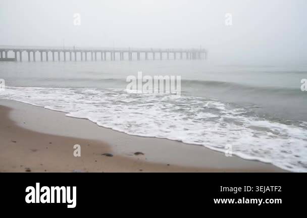 Calm sea waves softly lap sandy beach under dense fog with long pier ...