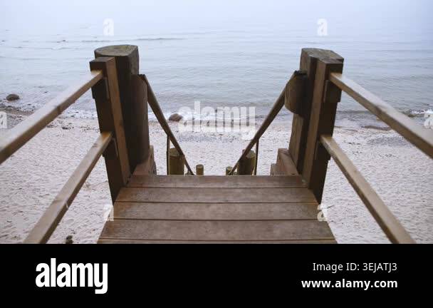 Rustic wooden steps with sturdy railings lead down to sandy beach with gentle waves under dense ...