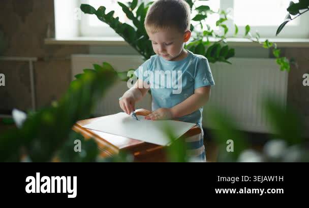 Cute toddler boy painting in a notebook surrounded by green houseplants ...
