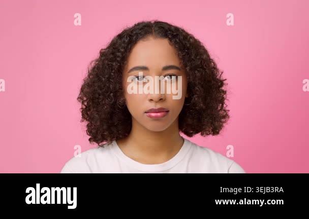 A young woman with curly hair stands confidently in front of a pink ...