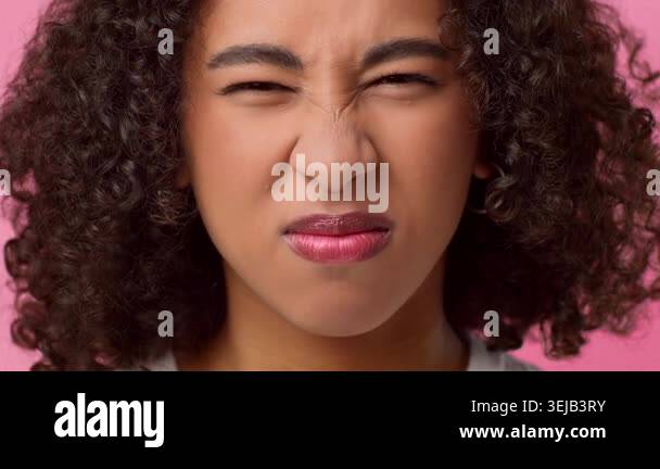 A young woman with curly hair stands in front of a pink background. She ...