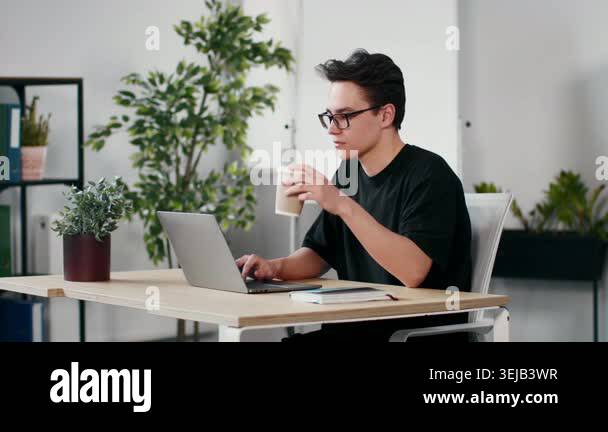 Young man with light skin and glasses is focused on his laptop at a ...
