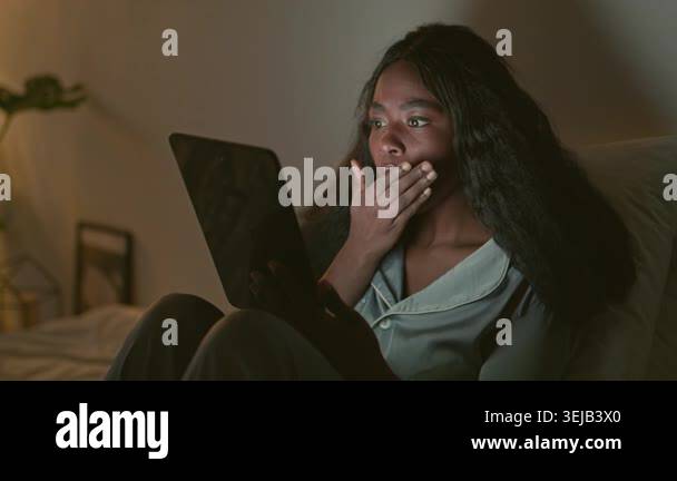 A woman with dark skin sits on her bed at home using a tablet. She ...
