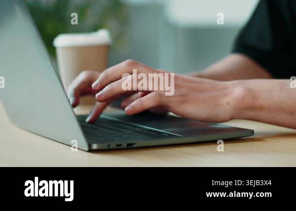 A person with light skin is typing on a laptop while sitting at a wooden table. A coffee cup is ...