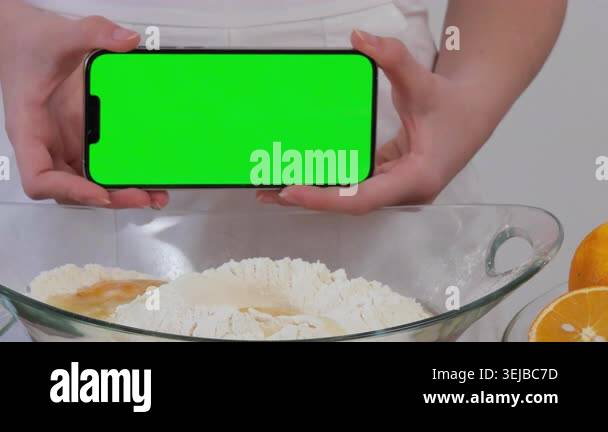 A young female baker holds a smartphone with a green screen while ...