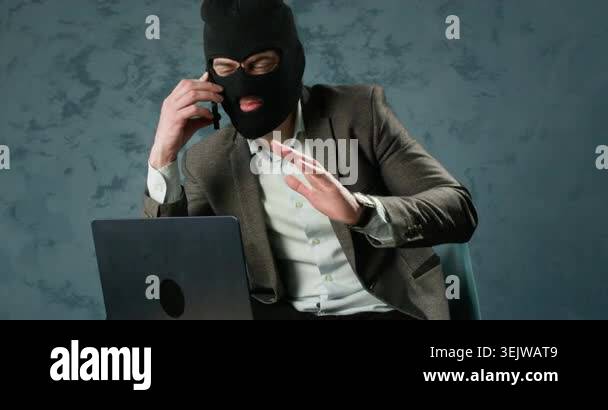 Scammer in balaclava talks on phone near laptop during online fraud ...
