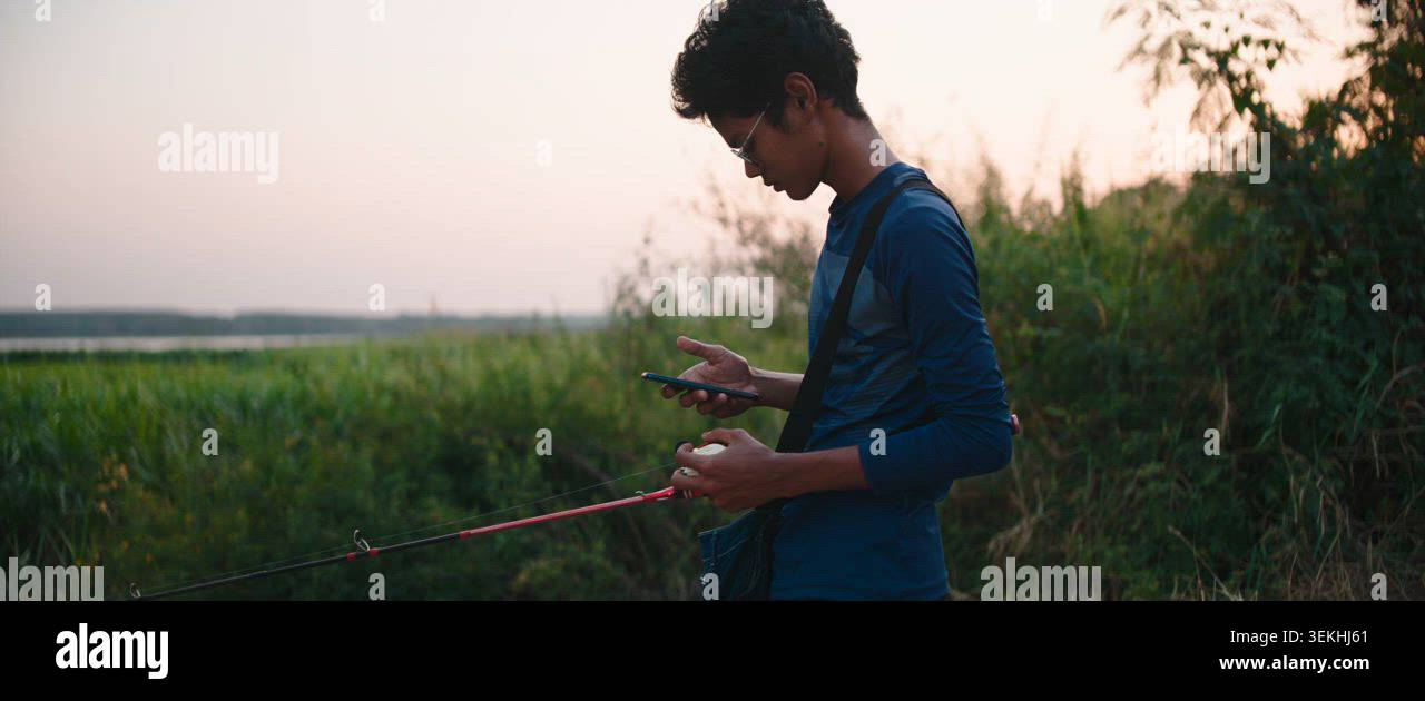Young fisherman using smartphone while holding a fishing rod in lake ...