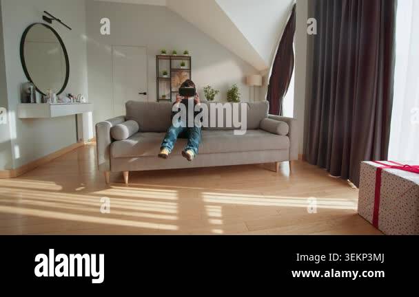 Young boy sits on a couch holding a virtual reality headset, ready for ...