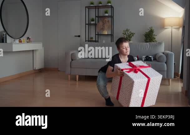 Young boy kneels on wooden floor, excitedly opening large, festive gift ...