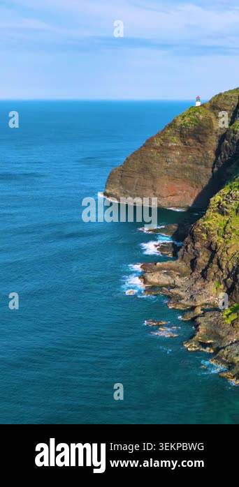Steep volcanic rock sea cliff with small lighthouse at summit, waves ...