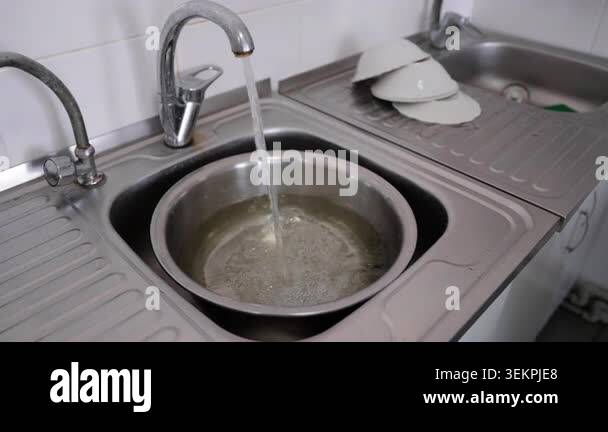 Water streams from a faucet into a bowl in a kitchen sink as a hand ...