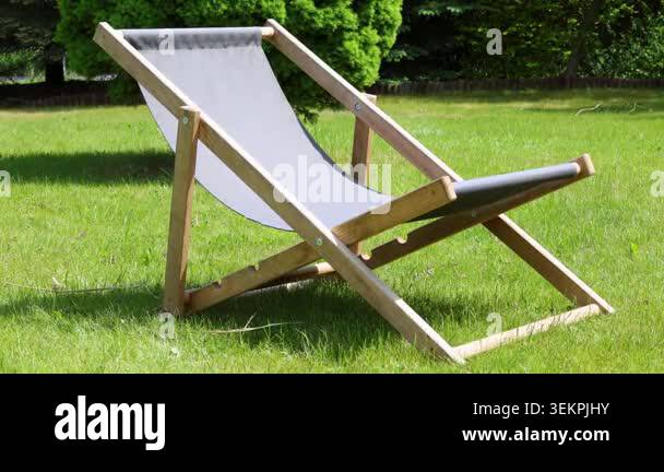 Simple wooden deck chair rests on green grass. Chair's fabric is light ...
