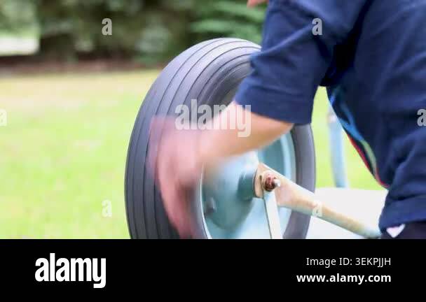 Child spins a wheelbarrow tire outdoors on a grassy lawn. A hand grips ...