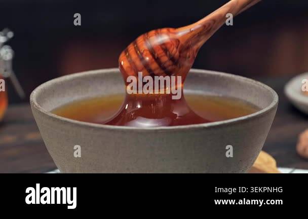 Golden honey flowing from wooden dipper into transparent glass bowl ...