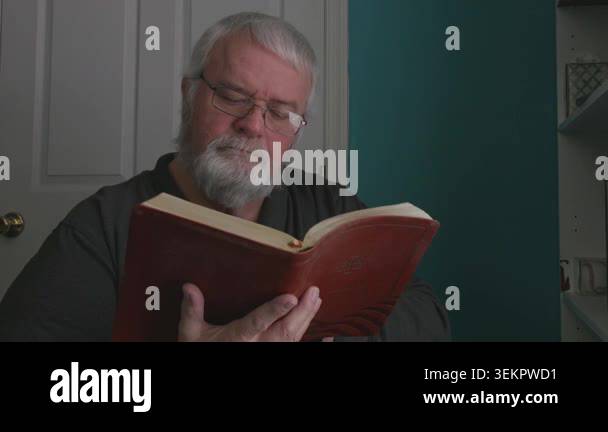 A man sits in a chair reading a book in a room with a blue wall. He ...