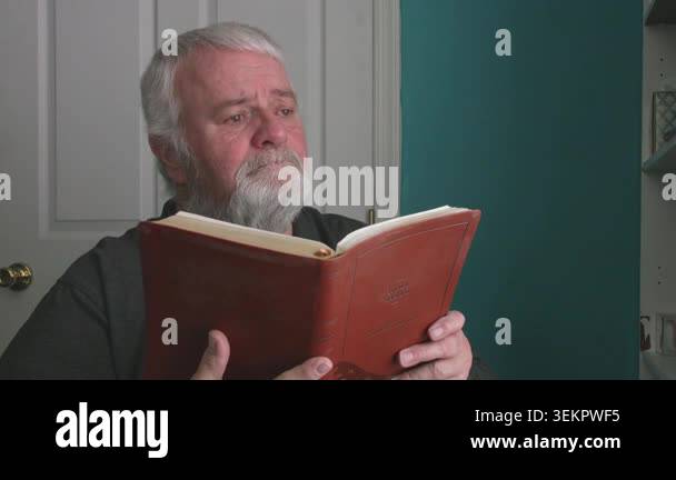 An older man sits in a quiet room looking at a book. He pauses often to ...