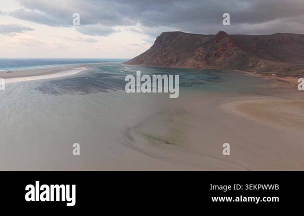 A wide coastal view with pale sand, calm shallow water, dark cliffs ...