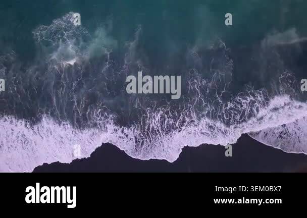 Top down aerial view of turquoise ocean waves crashing on a black sand ...