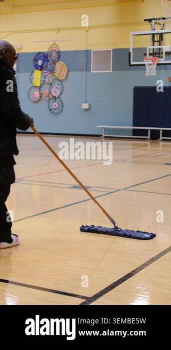 Maintenance worker pushing a dust mop to clean a shiny school gymnasium ...