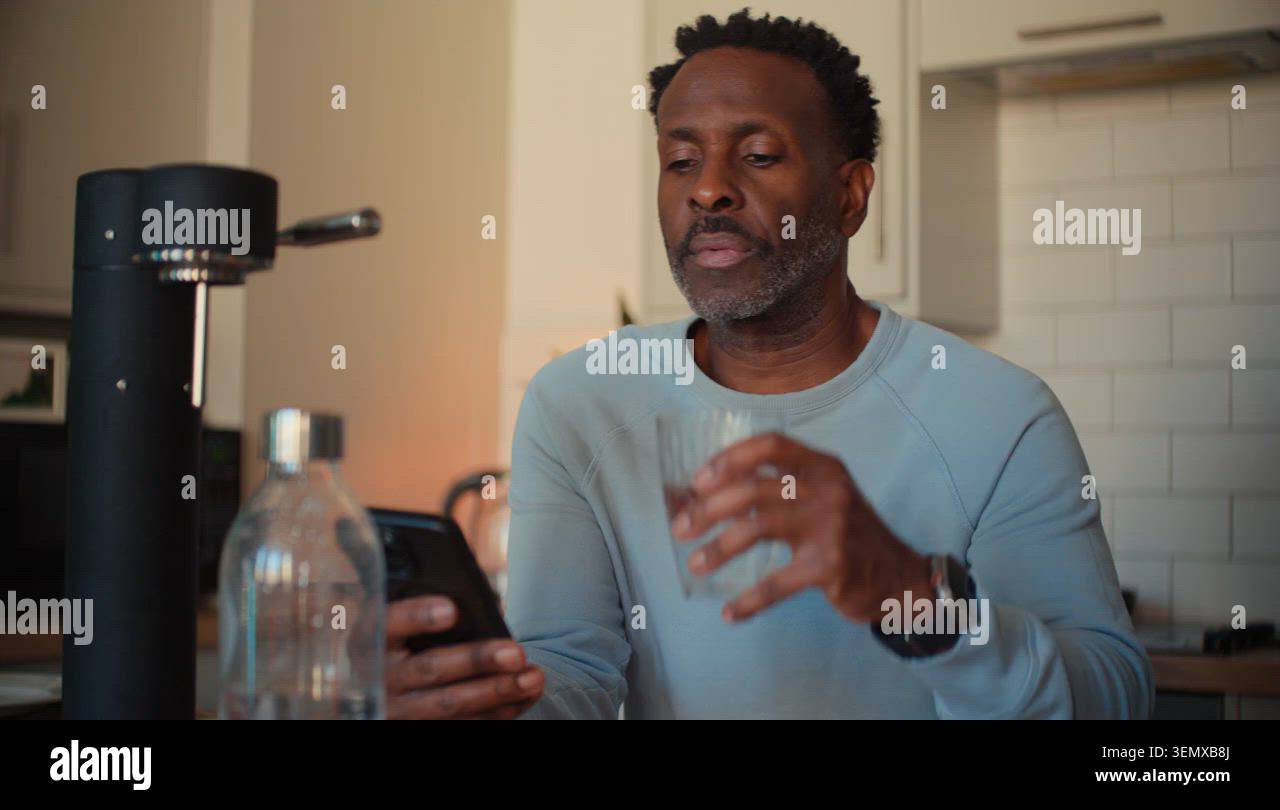 Man in Kitchen Checking Phone While drinking Glass of fizzy Water Stock ...