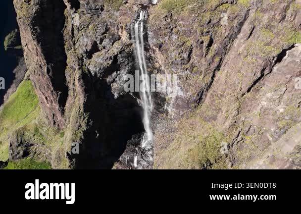 A tall narrow waterfall drops down a steep rocky cliff beside a dark ...