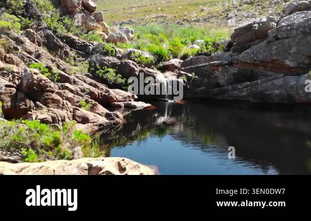 A small waterfall pours from a rocky hillside into a dark calm pool ...