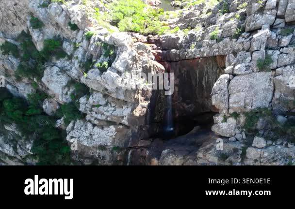 A thin waterfall flows through a rocky cliff opening into a dark pool ...