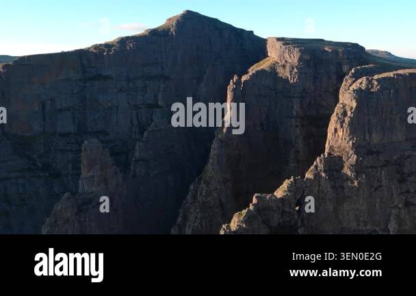 A dramatic rocky mountain cliff rises against a clear blue sky, with ...