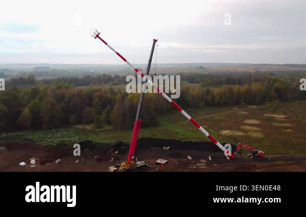A large crane raises a long red and white mast over a dirt worksite in ...